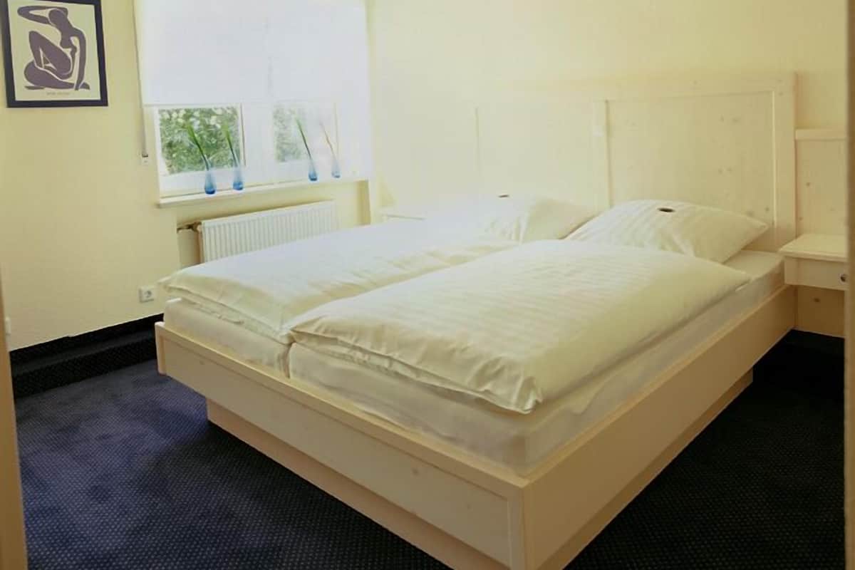 Cama de casal com roupa de cama branca em quarto com paredes em tom de bege, janela com persianas, radiador e quadro na parede.
