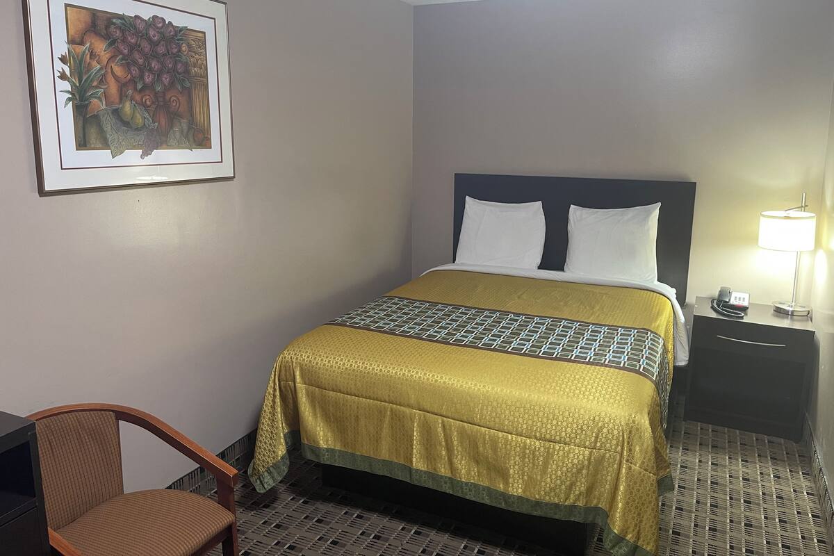 Quarto de hotel com uma cama, uma cadeira, uma mesa de cabeceira com abajur e um quadro na parede.