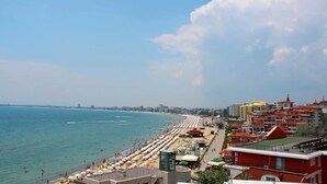 Uma praia com fileiras de guarda-sóis e espreguiçadeiras, um calçadão e edifícios com telhados vermelhos.