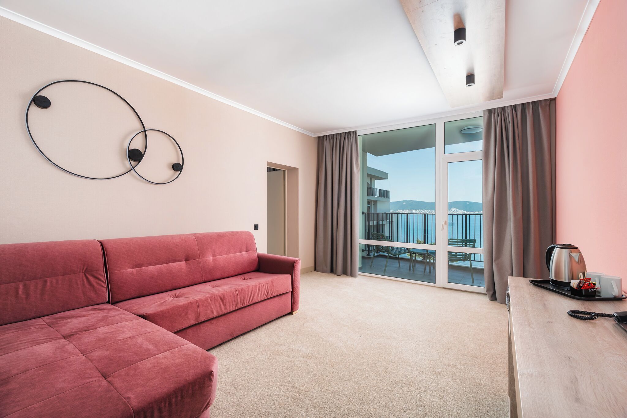 Quarto de hotel moderno com sofá vermelho, escrivaninha de madeira e grande janela com vista para a varanda e montanhas.