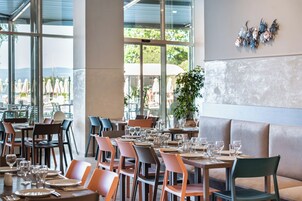 Um restaurante com mesas montadas para refeições, cadeiras e uma grande janela com vista para guarda-sóis externos.