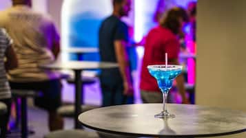 Uma área de bar com um cocktail azul sobre uma mesa redonda, rodeada por pessoas num espaço com iluminação roxa e pouca luz.