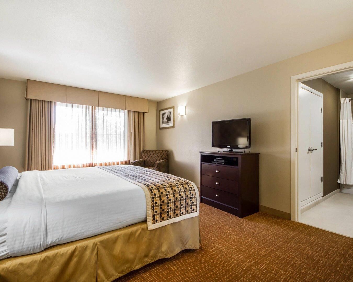 Quarto de hotel com uma cama grande, uma televisão em uma cômoda, uma cadeira e uma porta que dá para outro quarto.