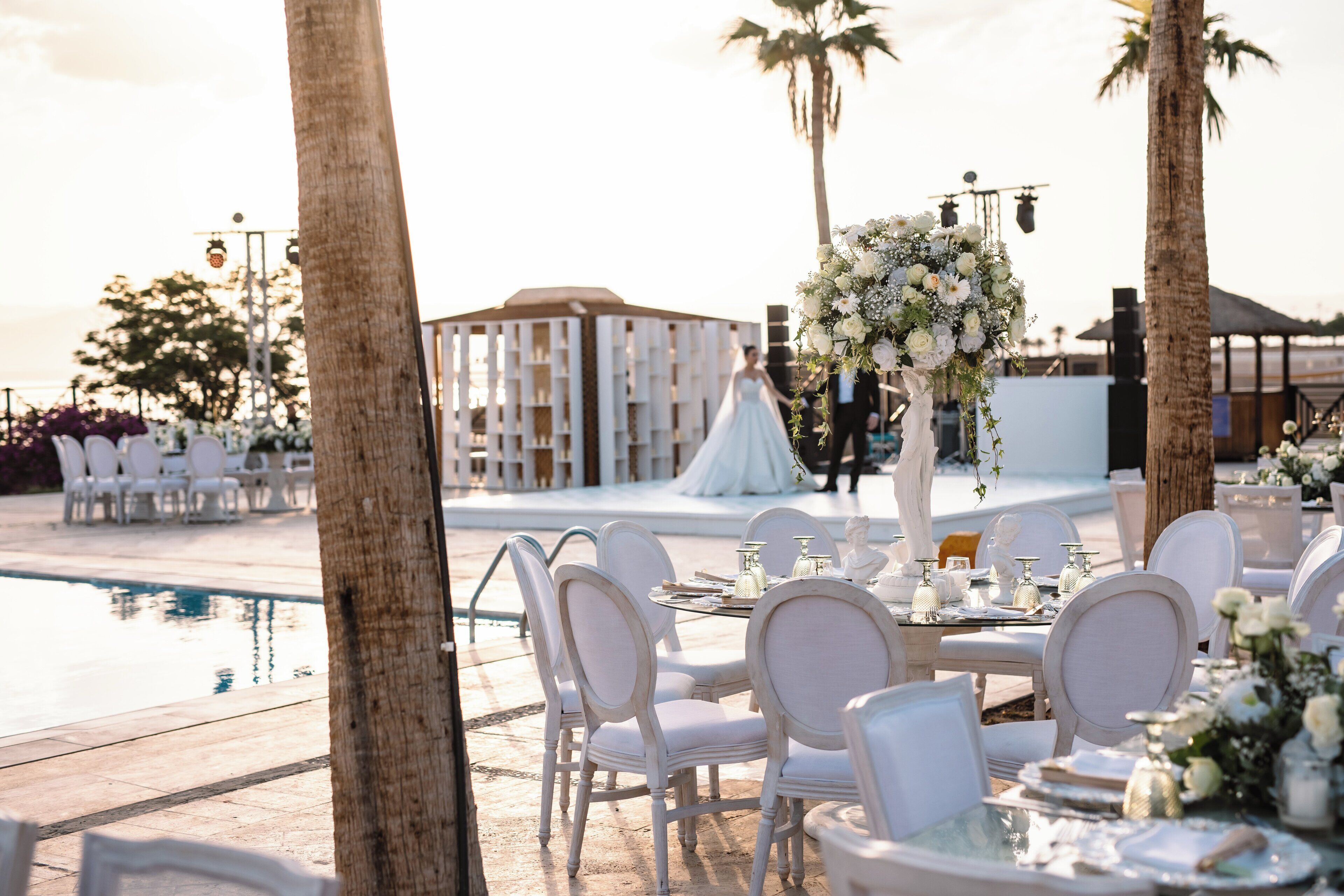 Decoração de casamento ao ar livre com cadeiras e mesas brancas, piscina e palmeiras.