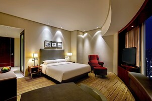 Quarto de hotel moderno com uma cama grande, uma poltrona vermelha, uma televisão e vista para a cidade.