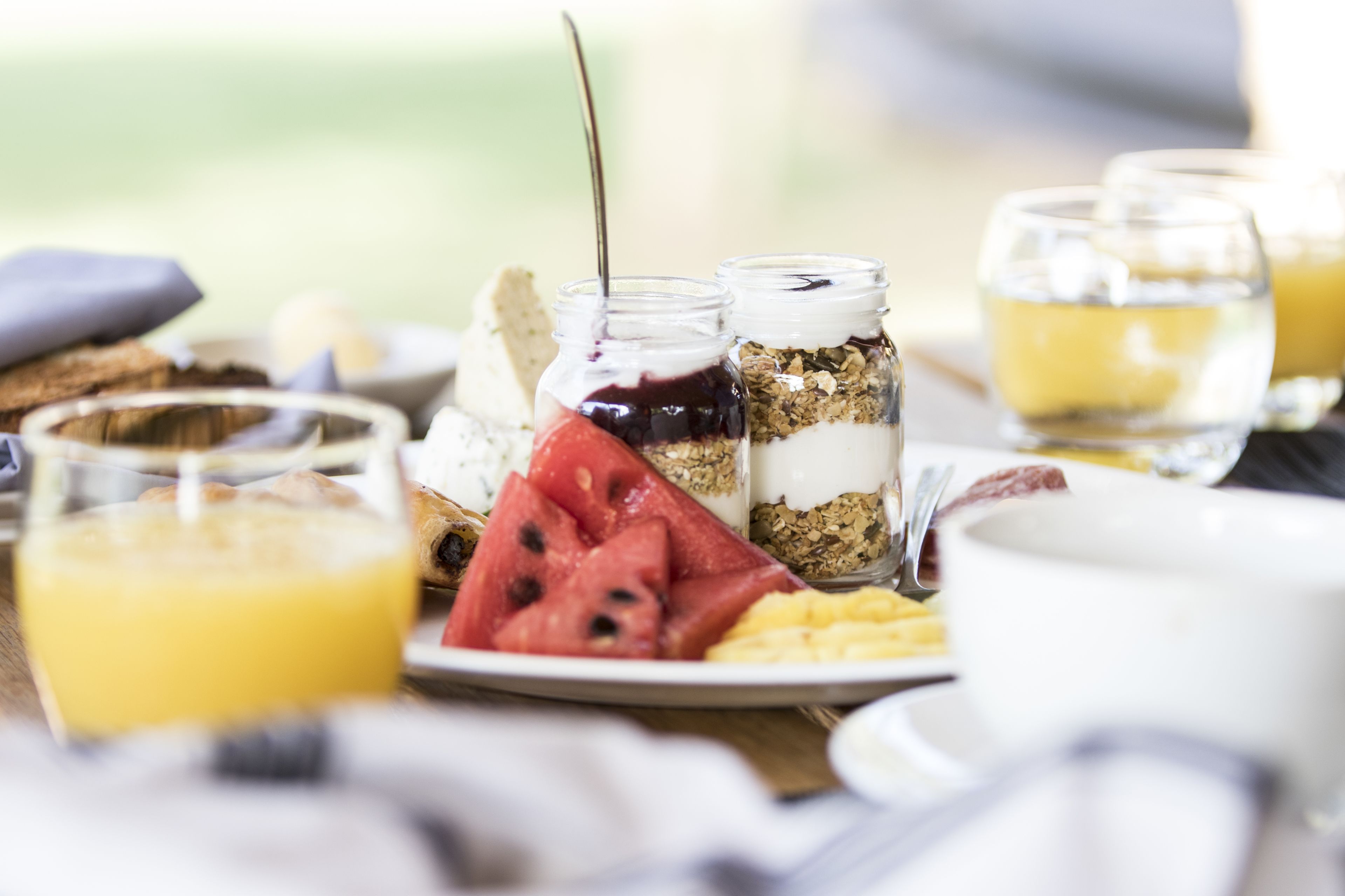 Uma mesa de café da manhã com diversos alimentos, incluindo parfaits de iogurte, granola, melancia e suco de laranja.