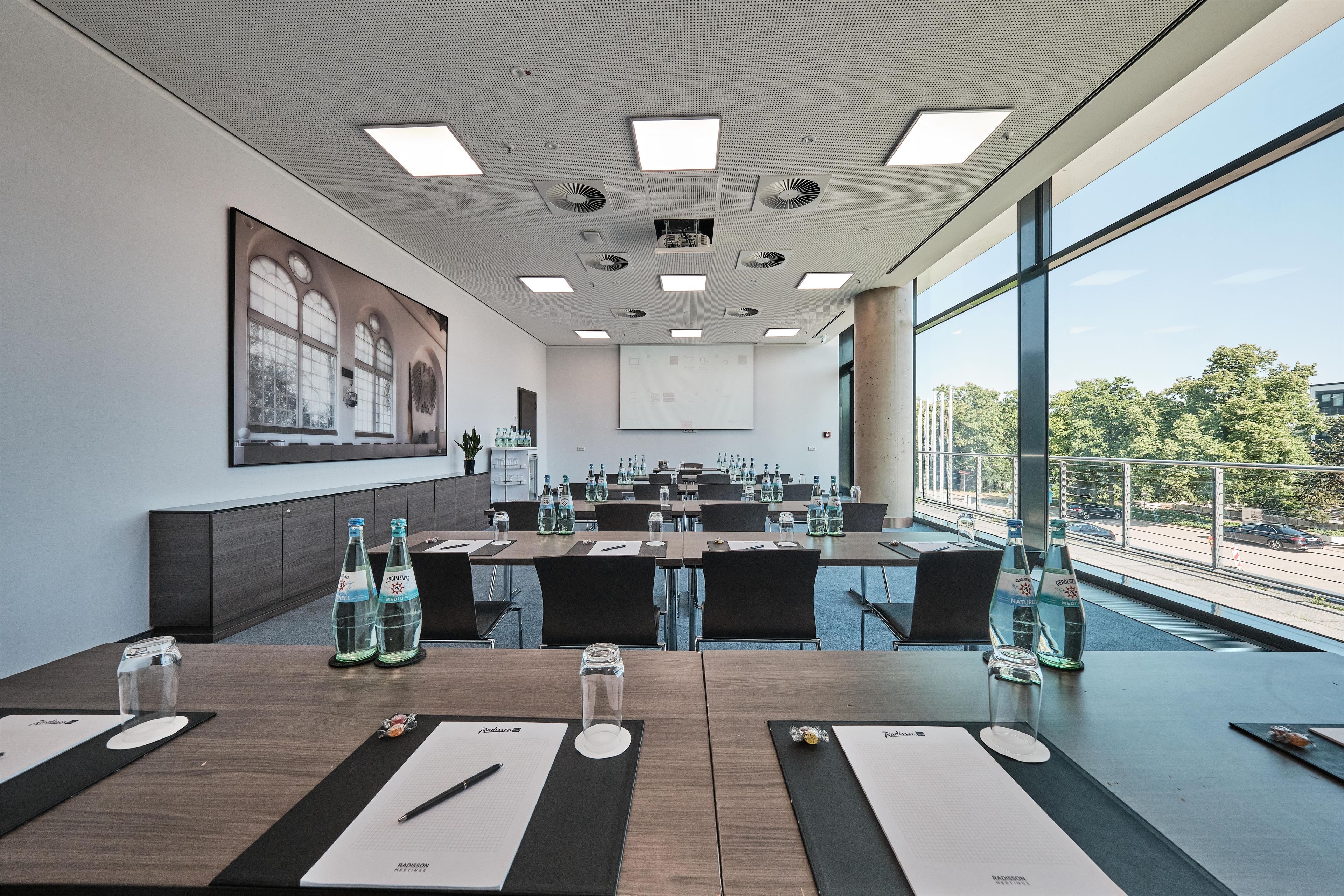 Uma sala de conferências com uma mesa grande, cadeiras, tela de projetor e vista para o exterior.