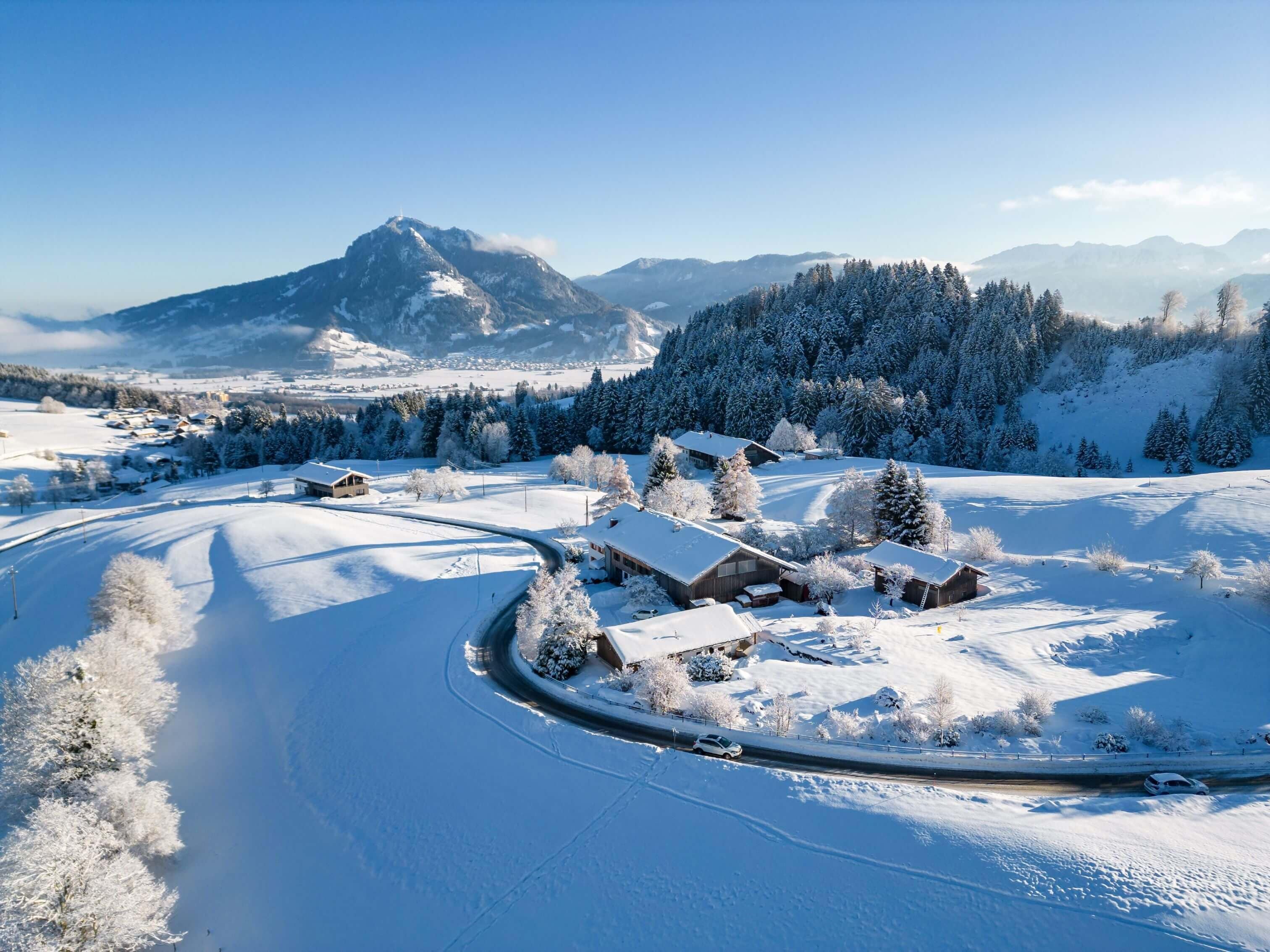 Paisagem coberta de neve com uma estrada sinuosa, vários edifícios e uma montanha ao fundo.