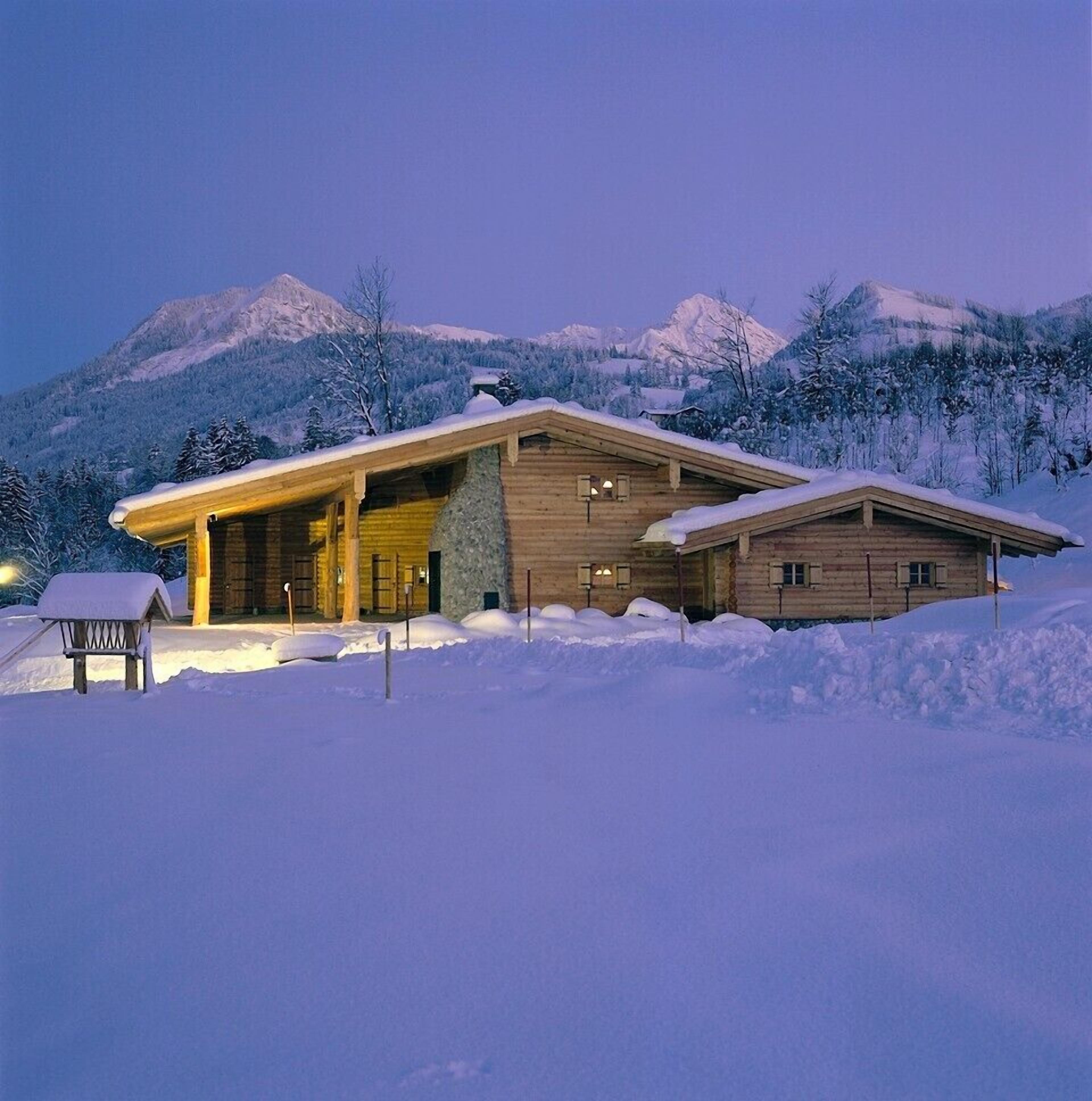 Uma cabana de madeira com varanda coberta, rodeada por neve e montanhas ao fundo.