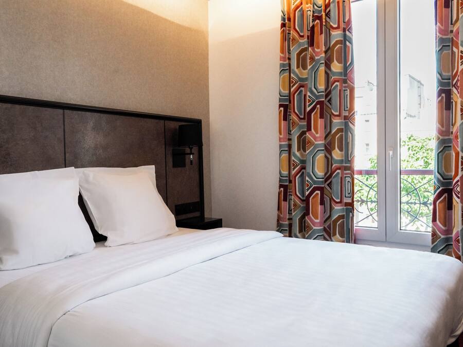Quarto de hotel com cama, mesa de cabeceira, abajur e janela com cortinas.