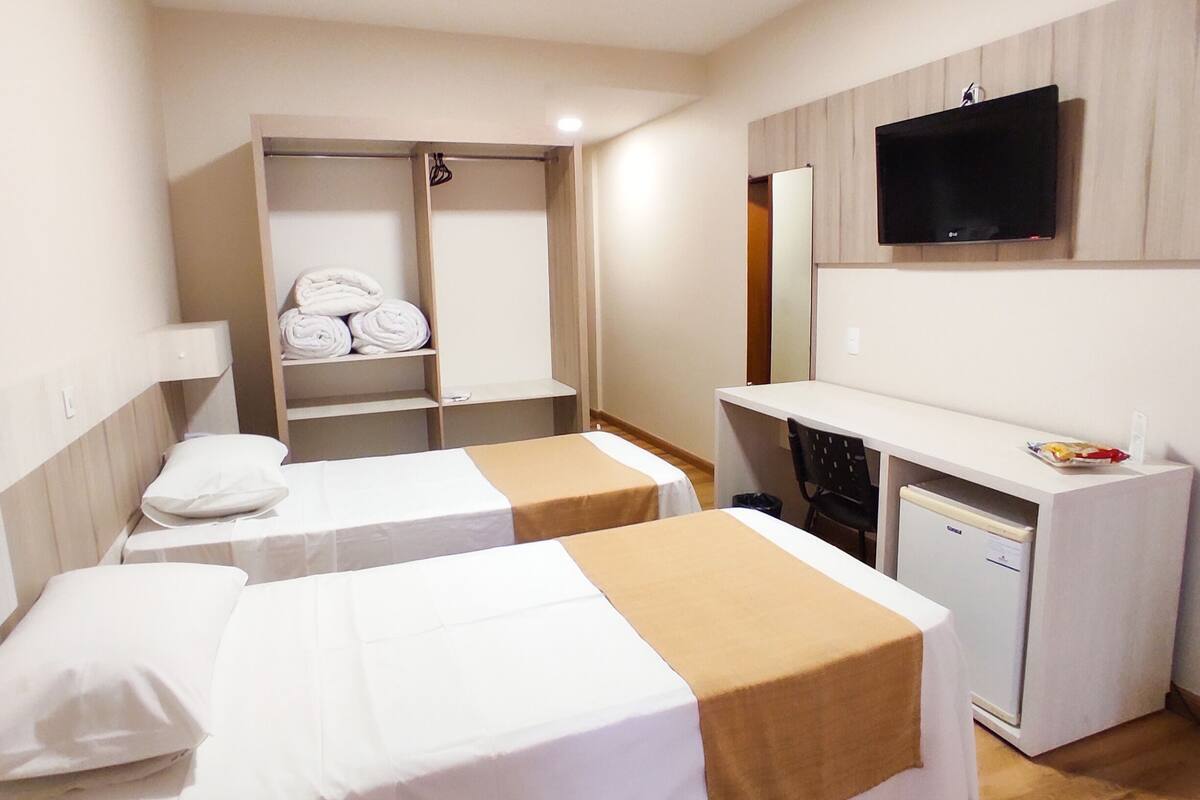 Quarto de hotel com duas camas, uma escrivaninha, uma televisão e um guarda-roupa.