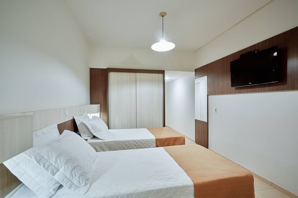 Quarto de hotel com duas camas, uma TV fixada na parede e uma luminária no teto.