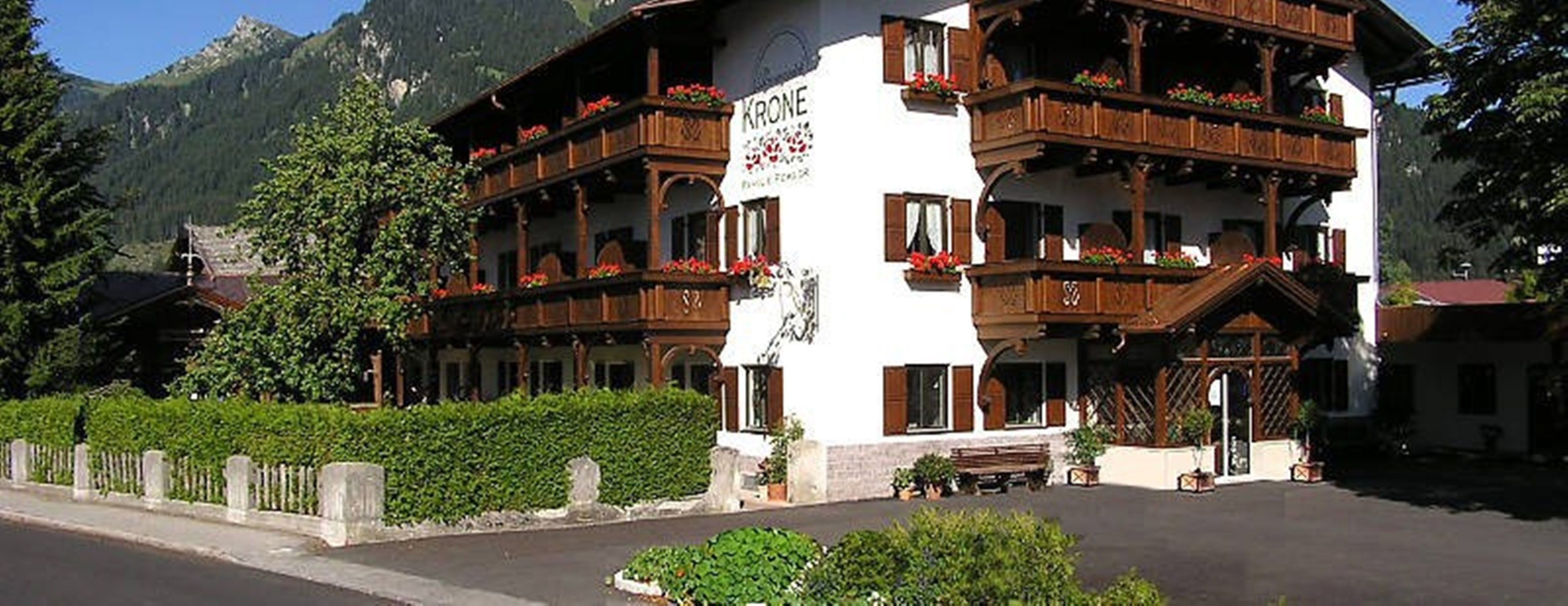 Um edifício tradicional de vários andares com varandas de madeira, uma placa com a inscrição "KRONE" e montanhas ao fundo.