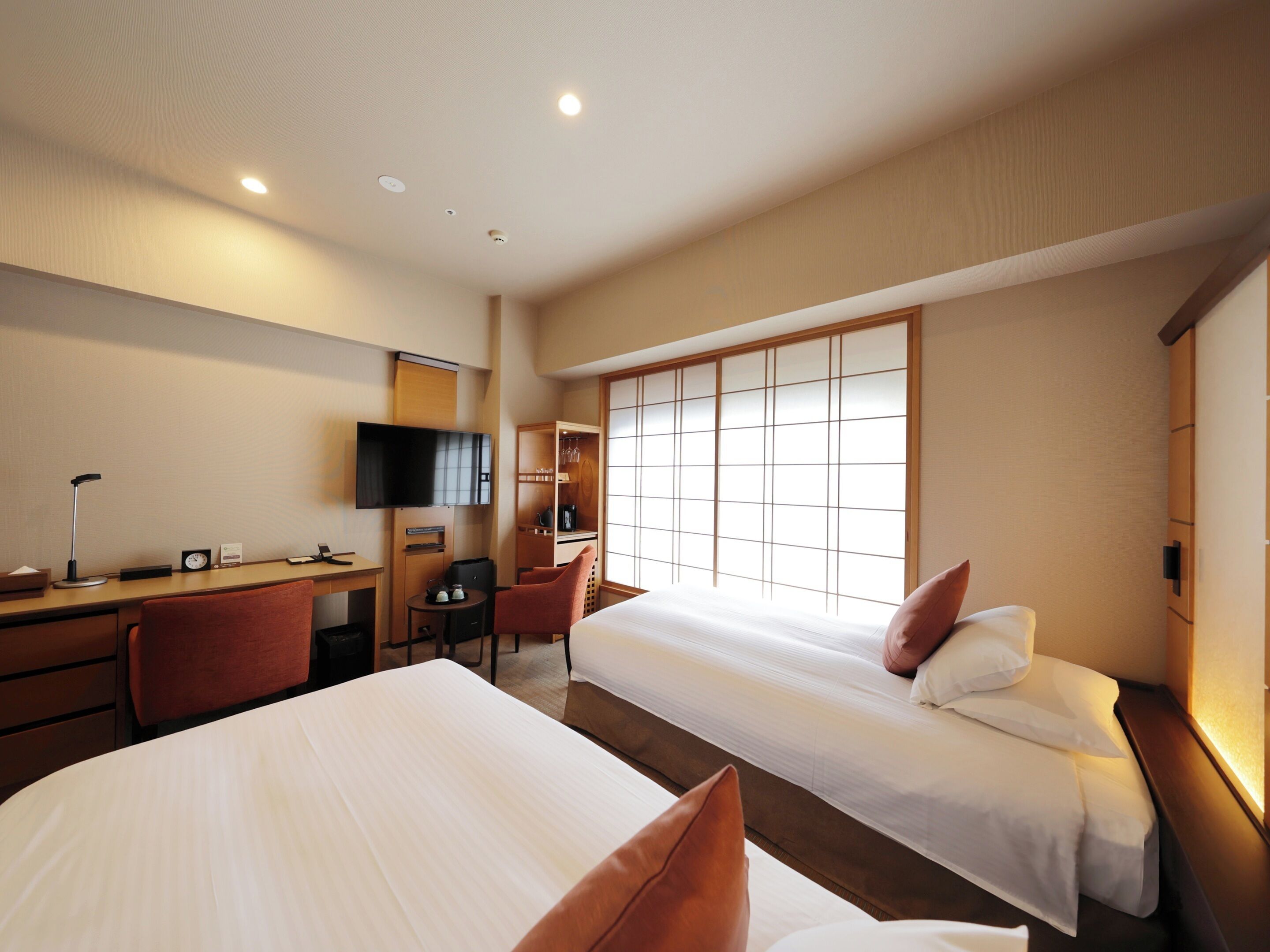 Quarto de hotel com duas camas, uma escrivaninha, uma cadeira, uma televisão e um guarda-roupa.