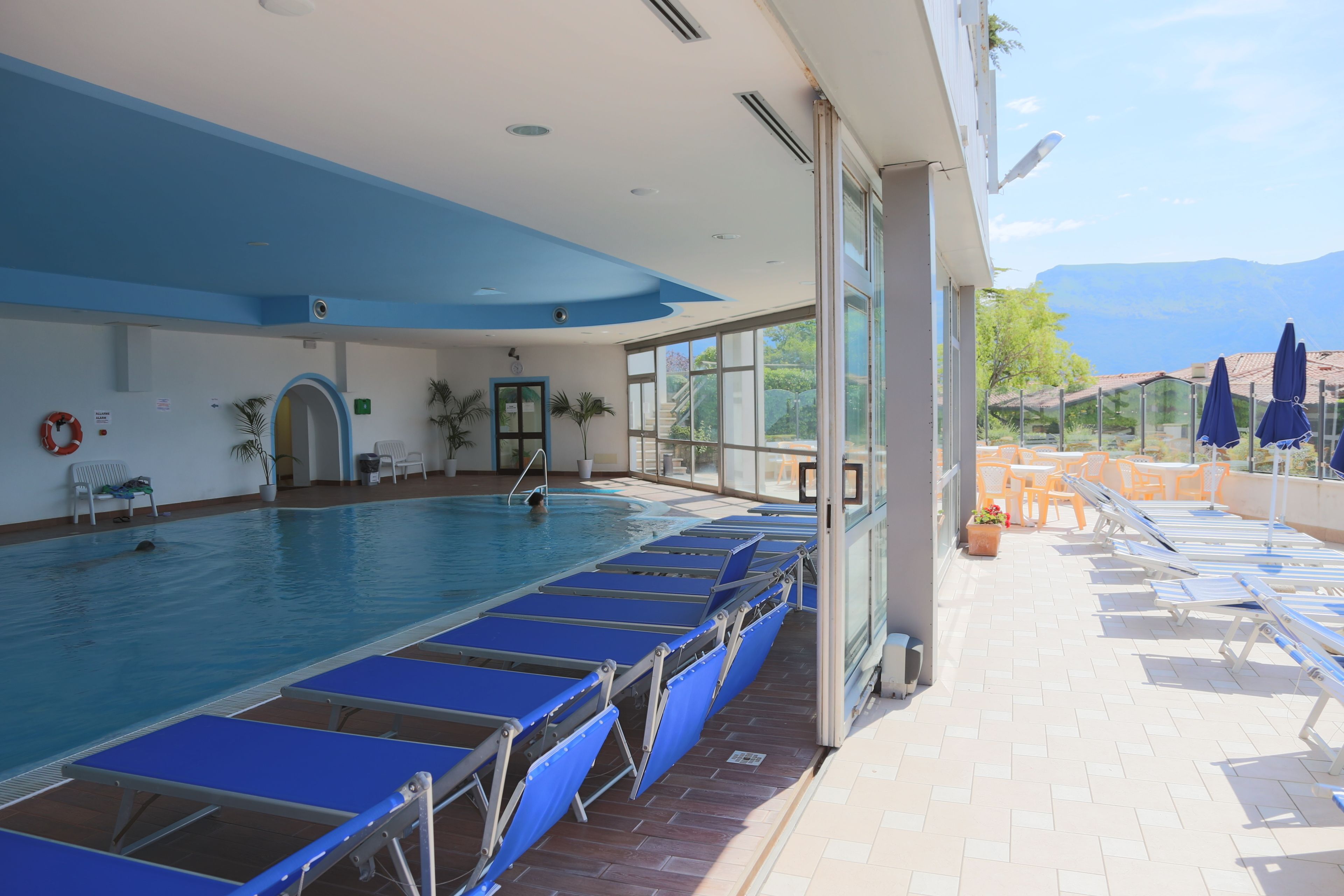 Piscina interior com espreguiçadeiras azuis, cadeira de salva-vidas e vista para as montanhas.