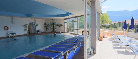 Piscina interior com espreguiçadeiras azuis, cadeira de salva-vidas e vista para as montanhas.
