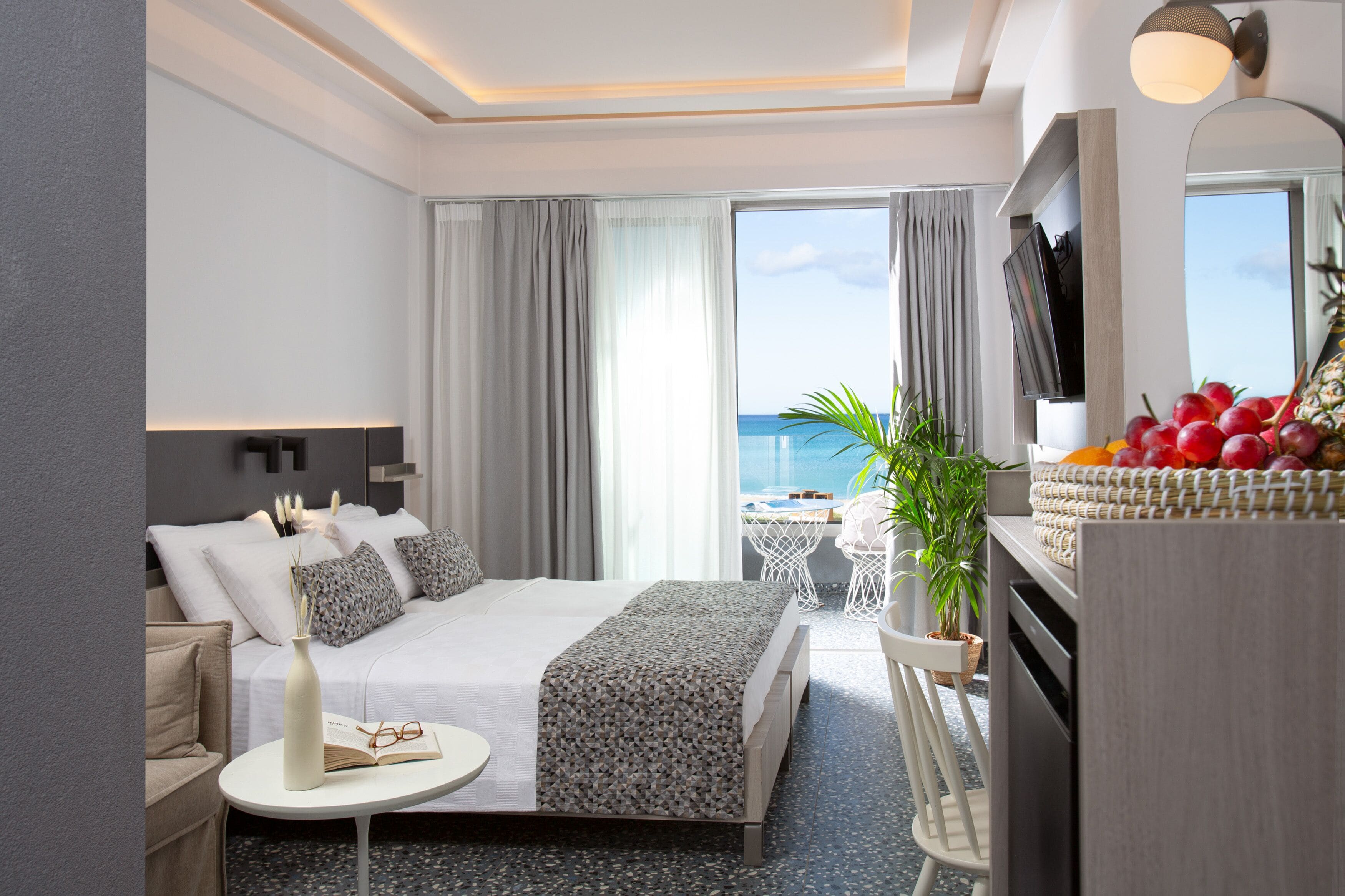 Quarto de hotel moderno com uma cama grande, uma mesa pequena com um vaso e um livro, uma televisão e uma varanda com vista para o mar.