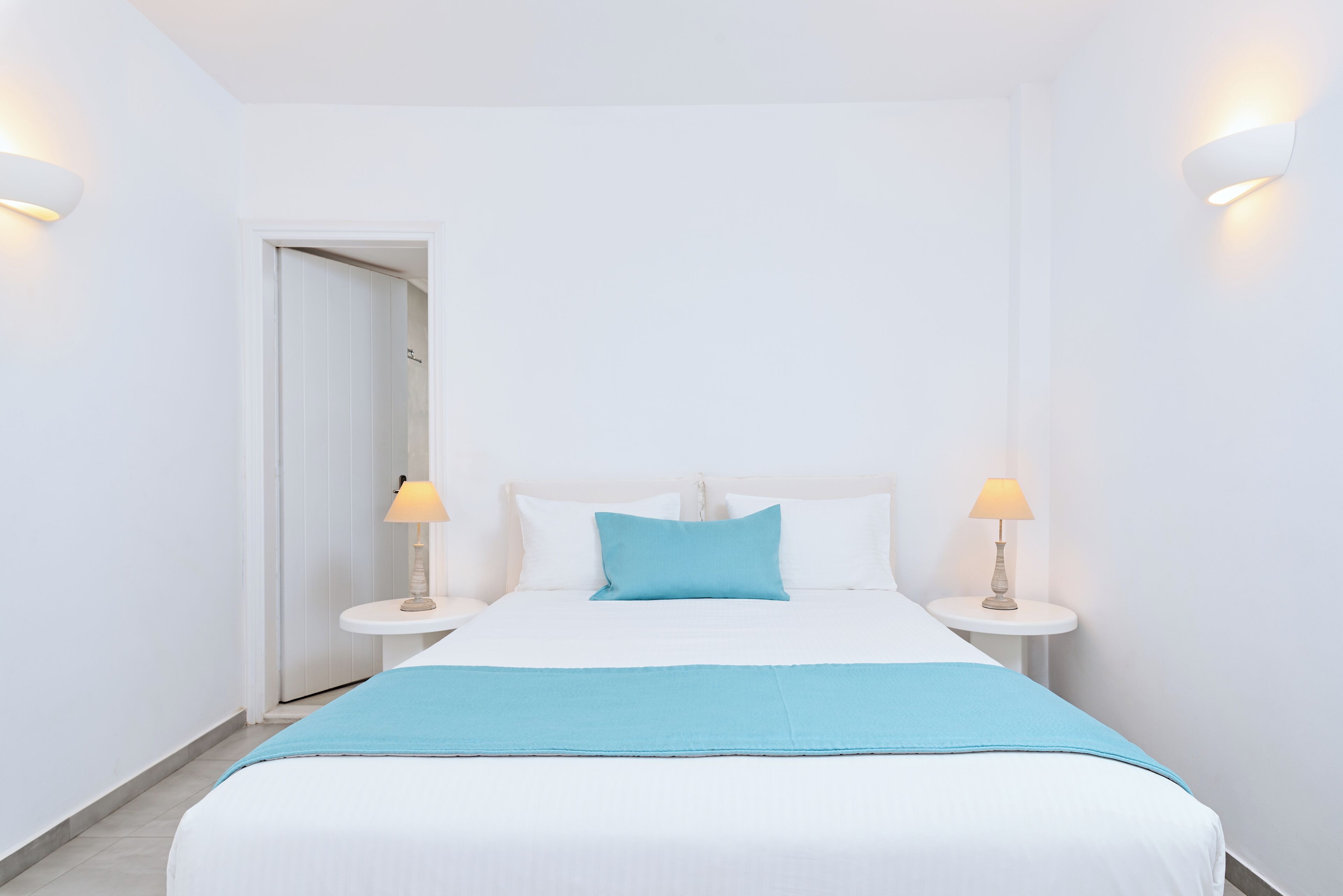 Quarto de hotel com uma cama, duas mesas de cabeceira com luminárias, uma porta e piso em tons claros.