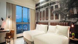 Quarto de hotel com duas camas, uma escrivaninha, uma cadeira, uma pequena mesa com uma planta e uma janela grande com vista para edifícios.