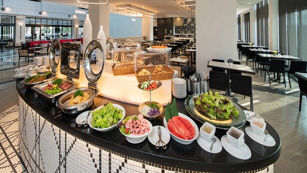 Uma área de buffet com diversas opções de comida, design moderno no teto e um amplo espaço de refeições com mesas e cadeiras.