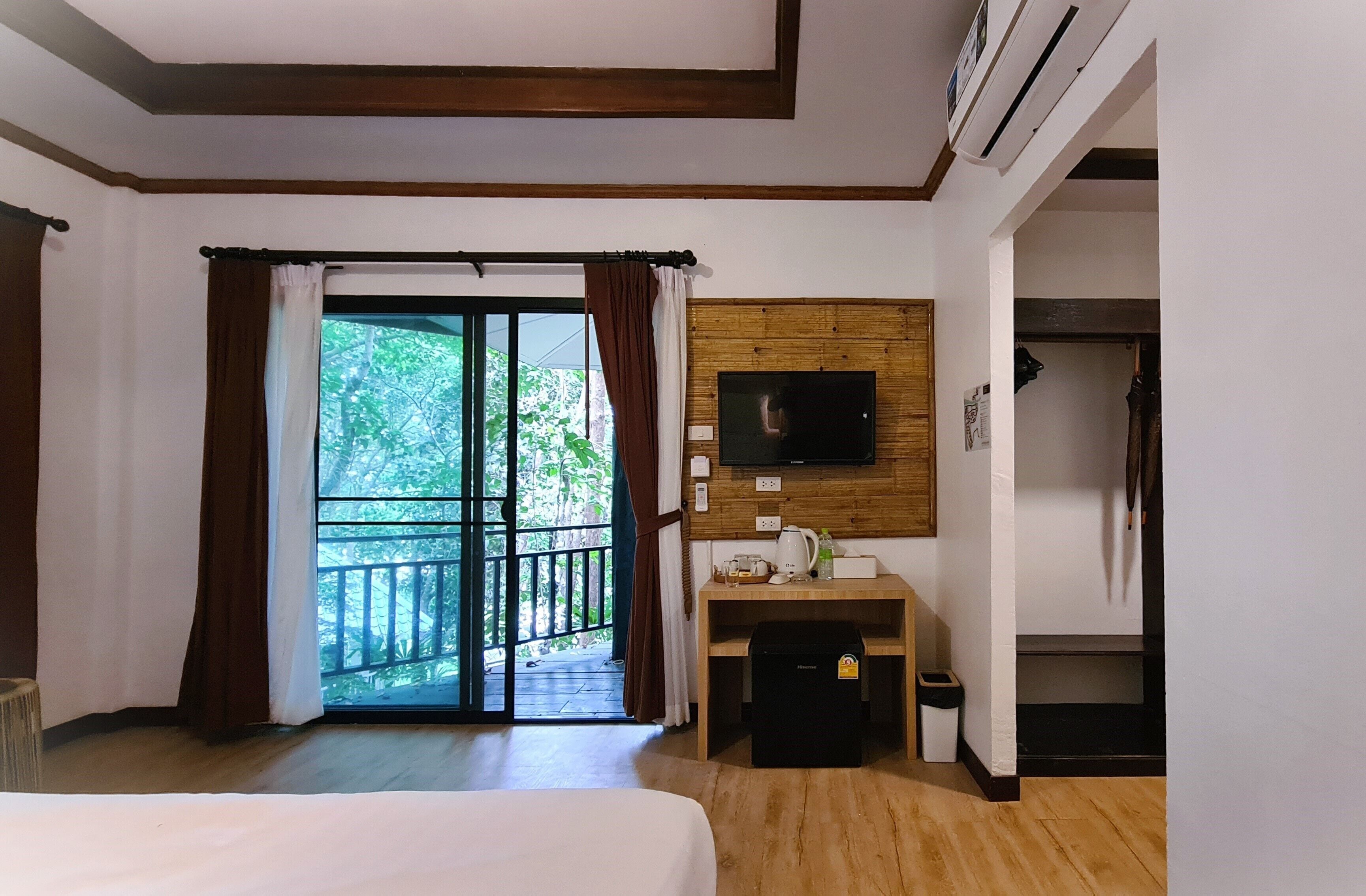 Quarto de hotel com cama, televisão fixada na parede, pequena mesa com chaleira e porta deslizante para uma varanda externa.