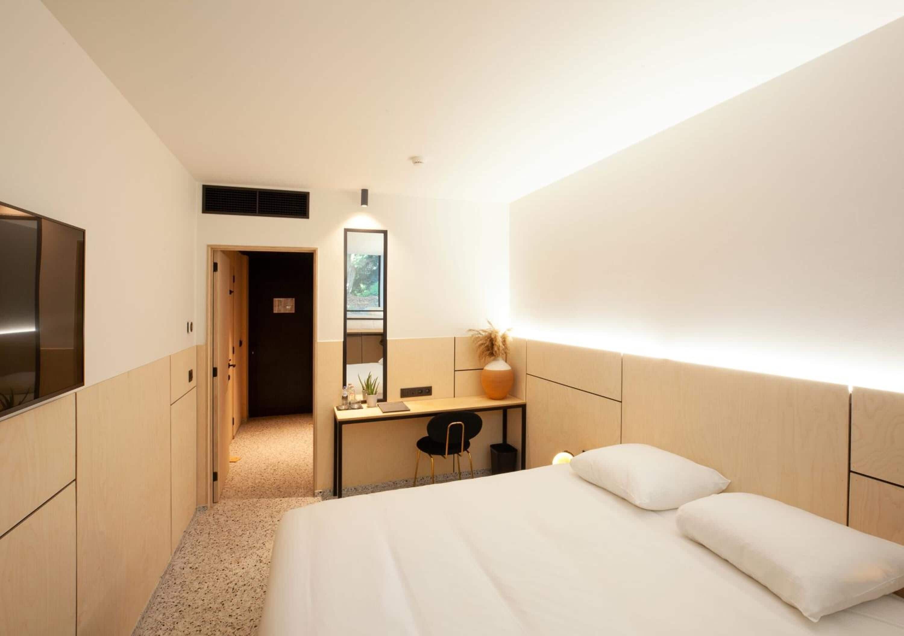 Quarto de hotel com cama, escrivaninha, cadeira, espelho e televisão fixada na parede.