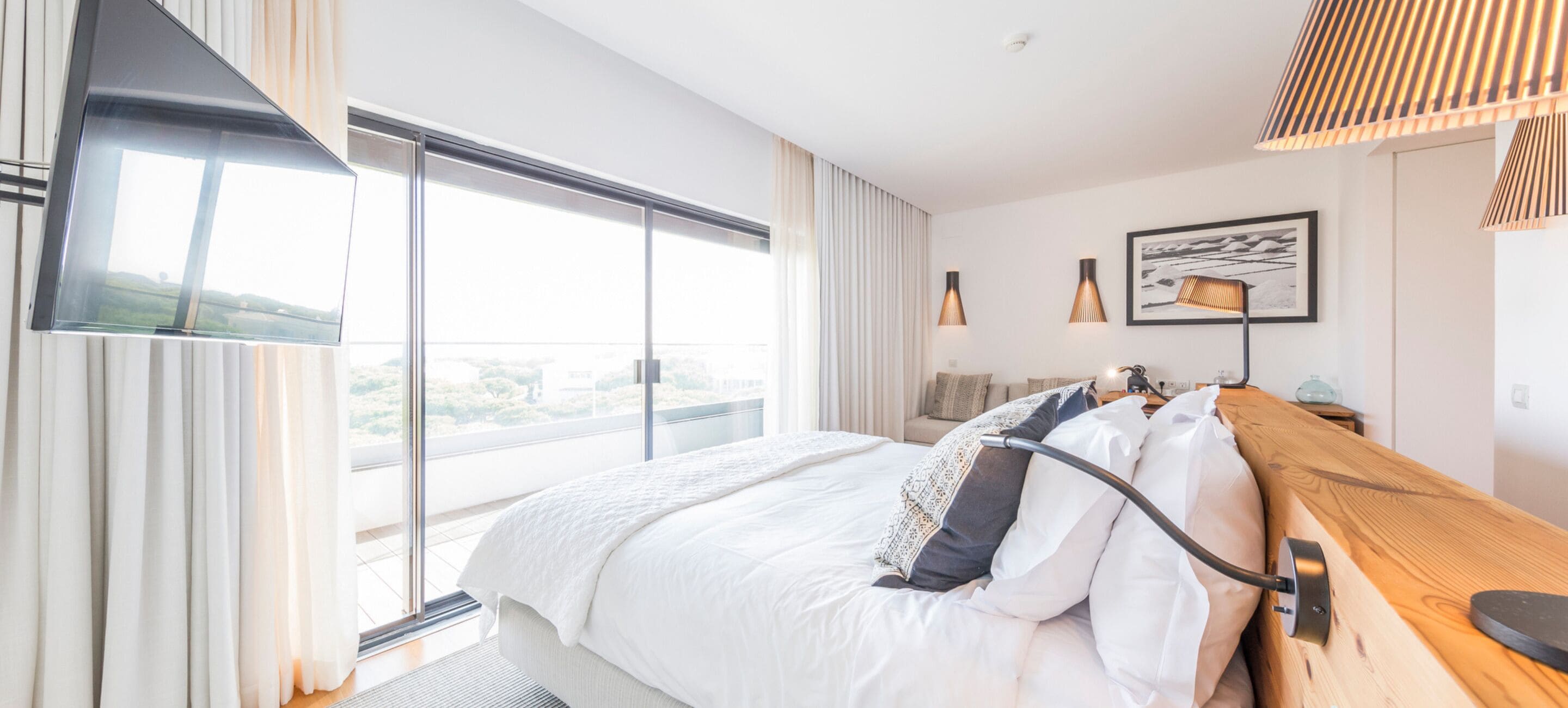 Quarto moderno com uma cama grande, cabeceira de madeira, mesa de cabeceira e vista da cidade através de janelas do chão ao teto.