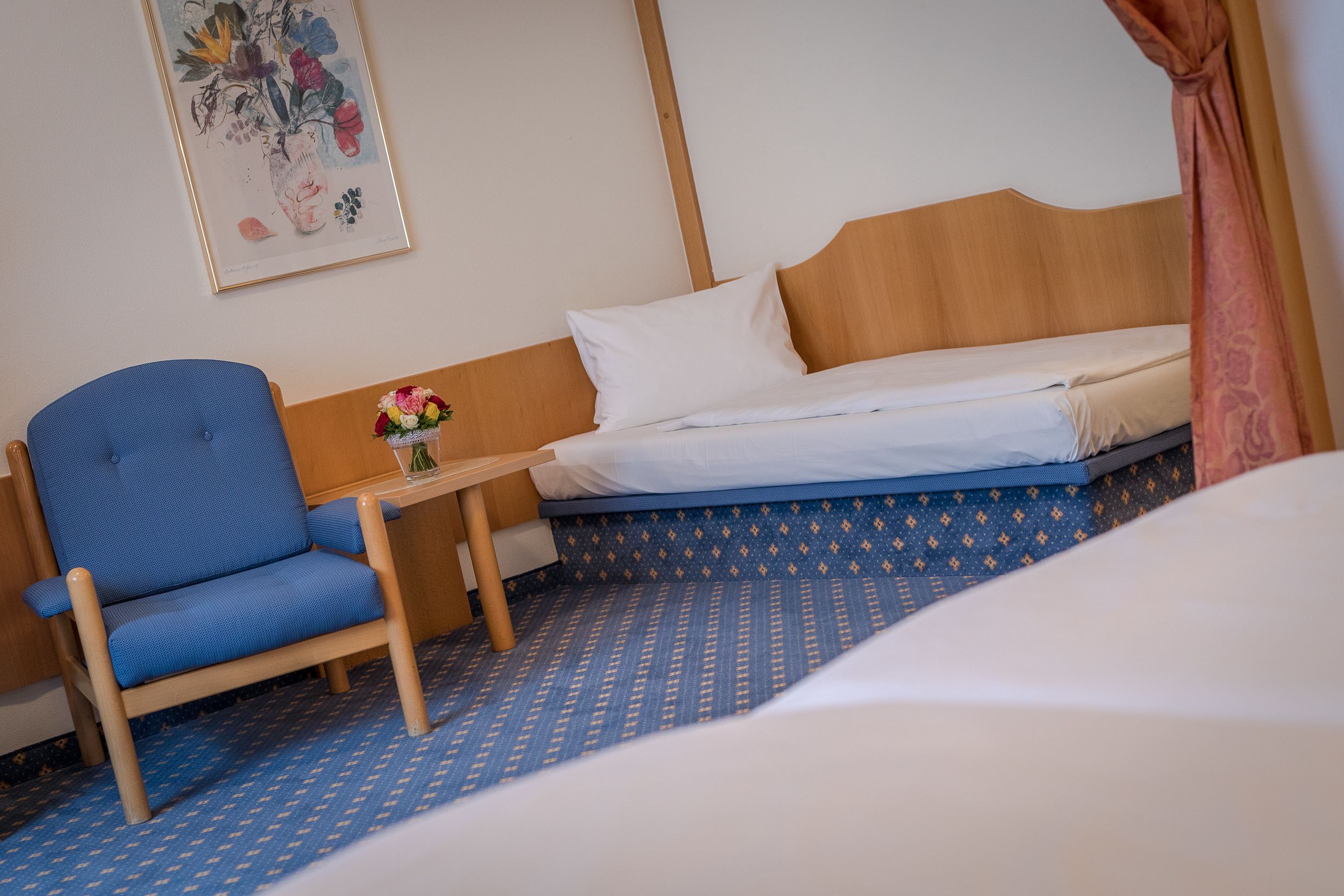 Quarto de hotel com uma cama de solteiro, uma cadeira azul, uma mesinha com um vaso de flores e um quadro pendurado na parede.