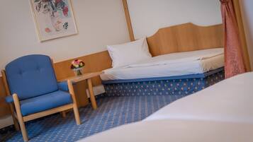 Quarto de hotel com uma cama de solteiro, uma cadeira azul, uma mesinha com um vaso de flores e um quadro pendurado na parede.