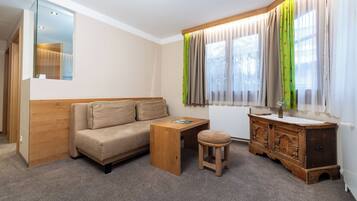 Quarto pequeno e aconchegante com sofá, mesa de madeira, um banquinho, armário de madeira e janela com cortinas leves.