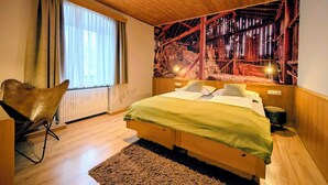 Quarto com teto de madeira, cama grande com manta verde, uma cadeira e um mural na parede com a imagem de um celeiro.