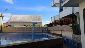 Uma piscina com uma fonte azul, um edifício moderno com telhado inclinado e um céu azul límpido.