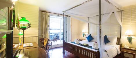 Uma cama com dossel com lençóis brancos, um criado-mudo de madeira com abajur, uma escrivaninha com abajur e uma cadeira.