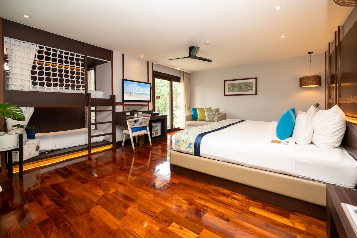 Quarto de hotel moderno com piso de madeira, cama com colcha azul e branca, escrivaninha com cadeira, televisão e uma janela grande.