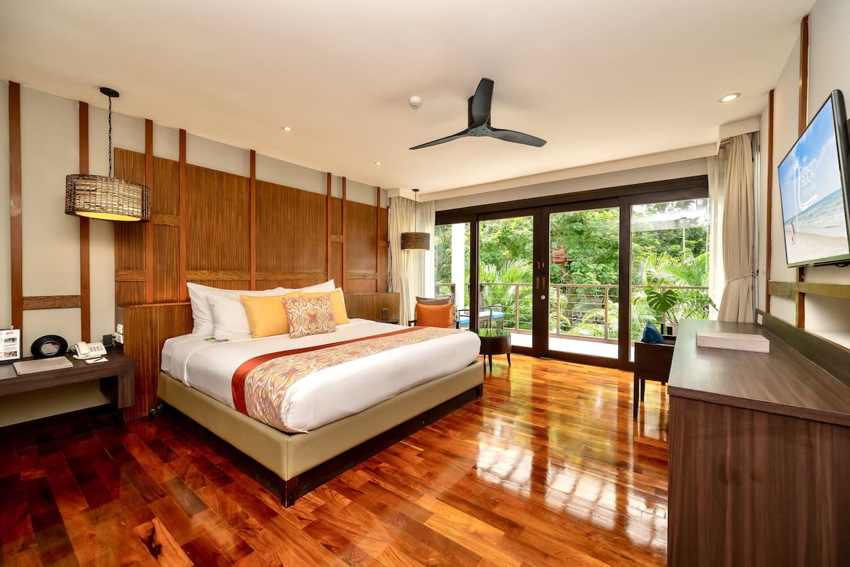 Um quarto espaçoso com uma cama grande, painéis de madeira, ventilador de teto e vista para o verde através de amplas janelas.