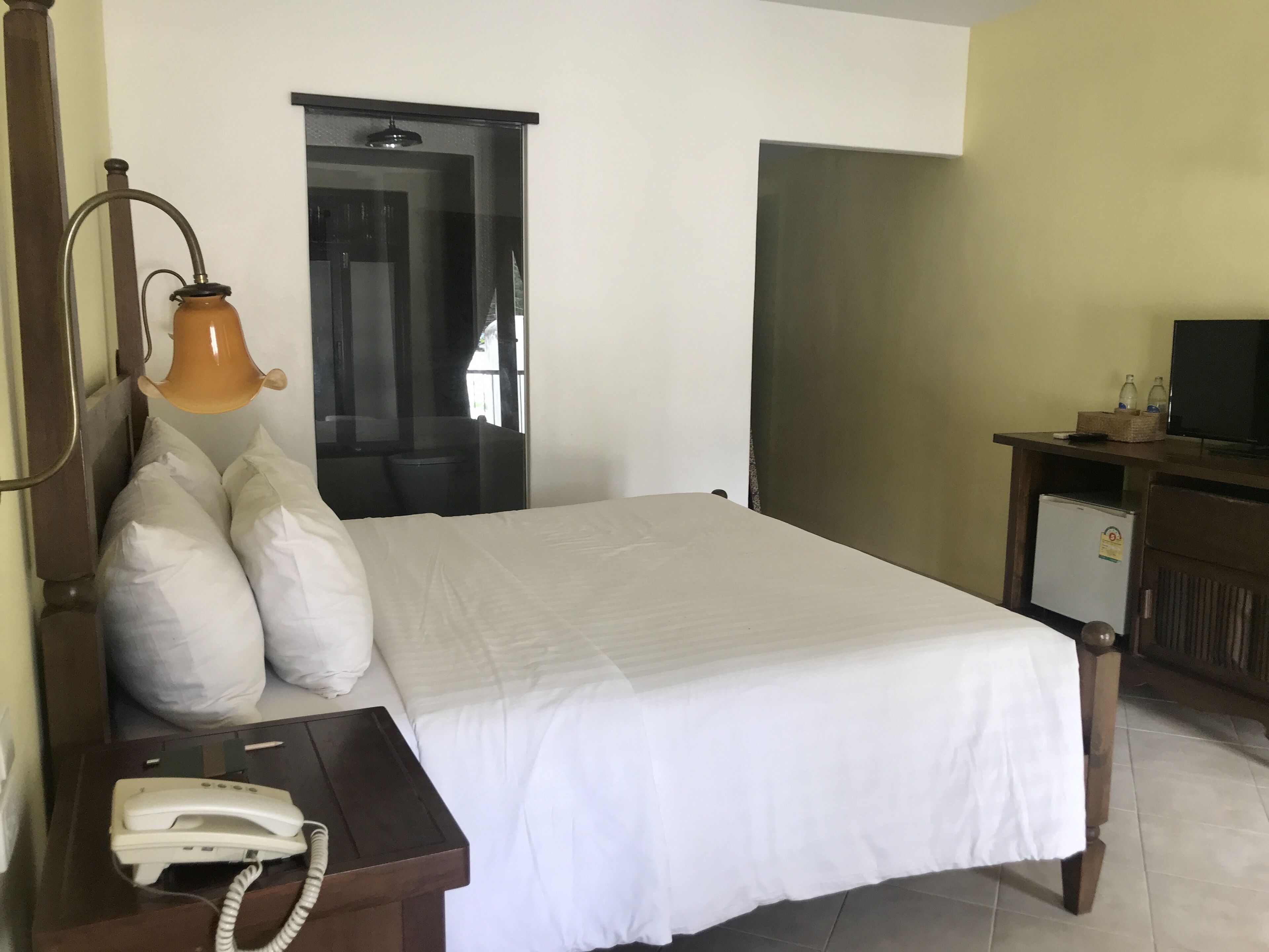 Quarto de hotel com cama, telefone de apoio, luminária, televisão e pequeno frigorífico.