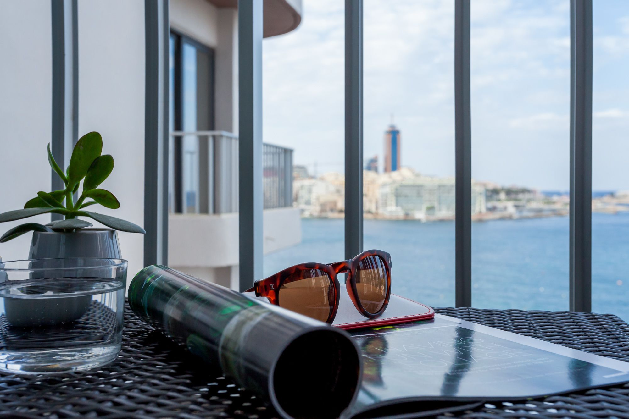 Uma mesa com uma planta, um objeto enrolado e óculos de sol, com vista para um horizonte urbano com água e edifícios.
