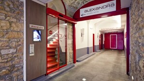 Entrada de hotel com parede de pedra, placa vermelha e branca para 'Alexandra Hotel' e escadaria que leva aos andares superiores.