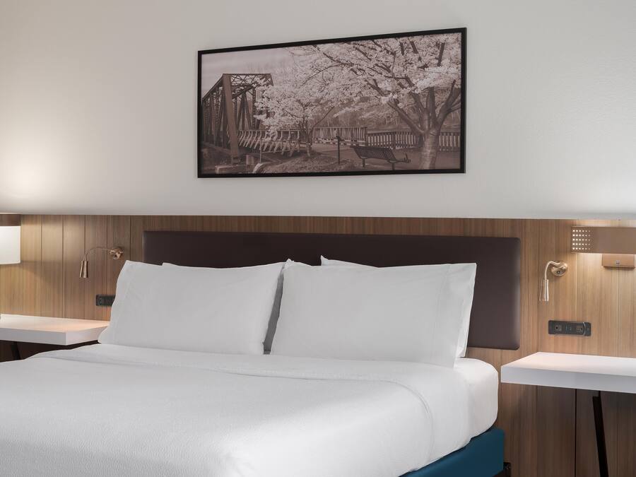 Quarto de hotel com cama, mesa de cabeceira, luminária na parede e um quadro emoldurado sobre a cama.