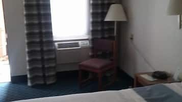 Quarto de hotel com cama, uma cadeira, escrivaninha, luminária e janela com cortinas.