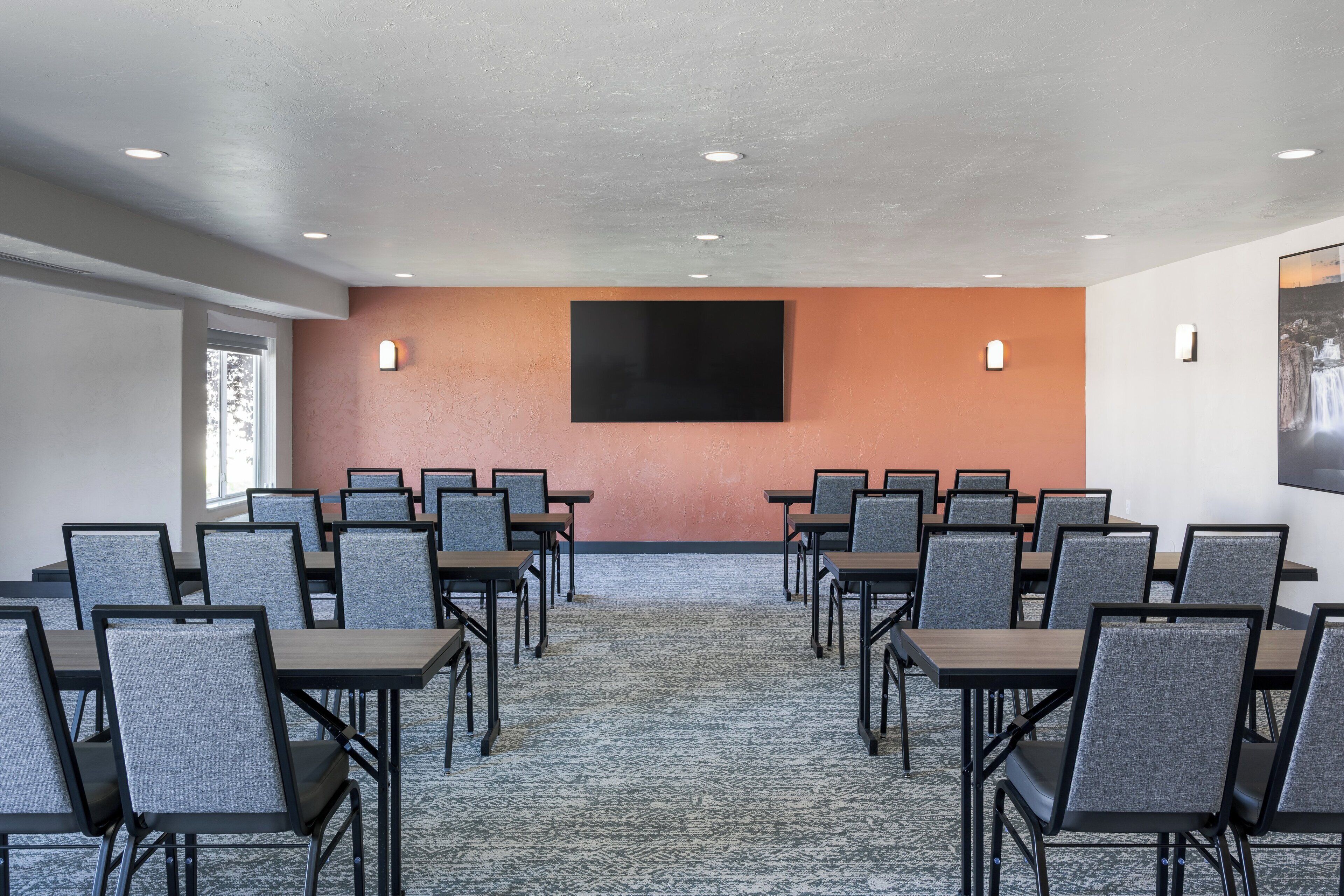 Uma sala de conferências com fileiras de cadeiras e mesas, uma televisão de tela plana grande na parede e uma janela no lado esquerdo.