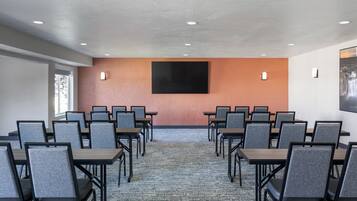 Uma sala de conferências com fileiras de cadeiras e mesas, uma televisão de tela plana grande na parede e uma janela no lado esquerdo.