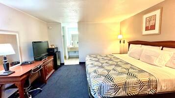 Quarto de hotel com cama, escrivaninha com televisão, pequeno frigorífico e casa de banho visível através de uma porta aberta.