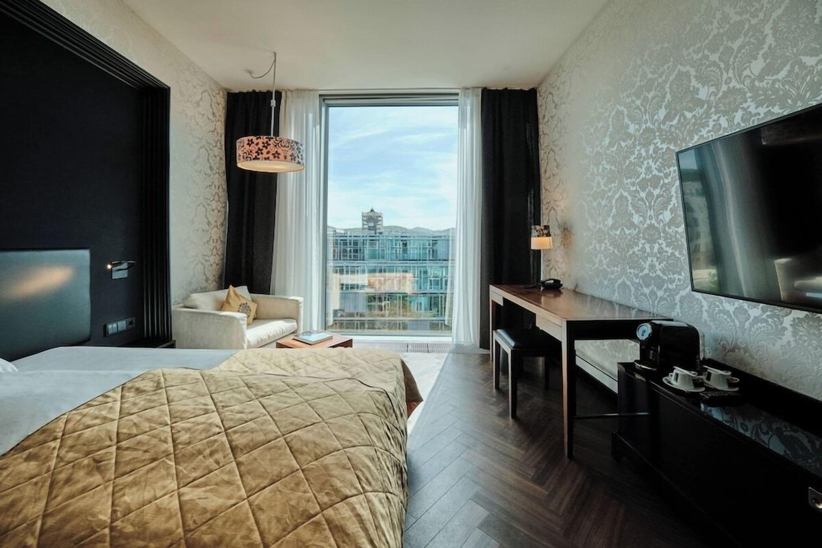 Quarto de hotel com cama grande, escrivaninha, cadeira, televisão e vista para a paisagem urbana.
