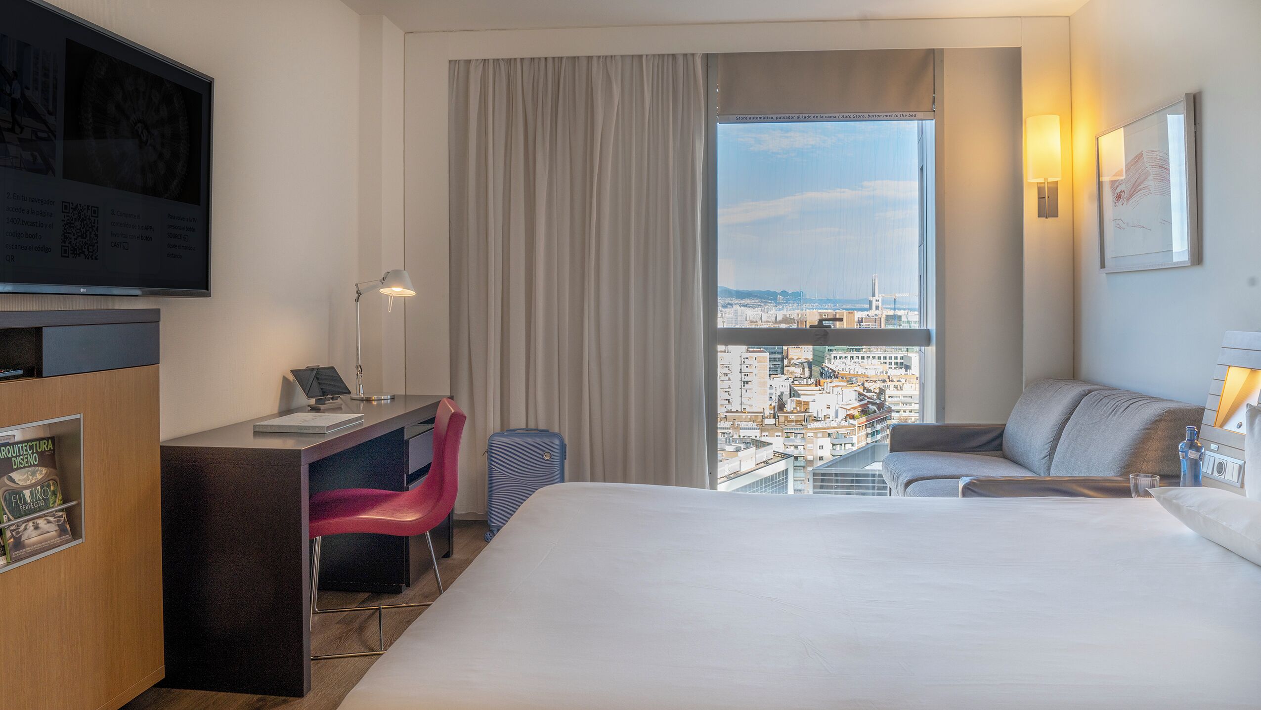 Quarto de hotel com cama, uma escrivaninha com cadeira, televisão e uma janela grande com vista para a cidade.