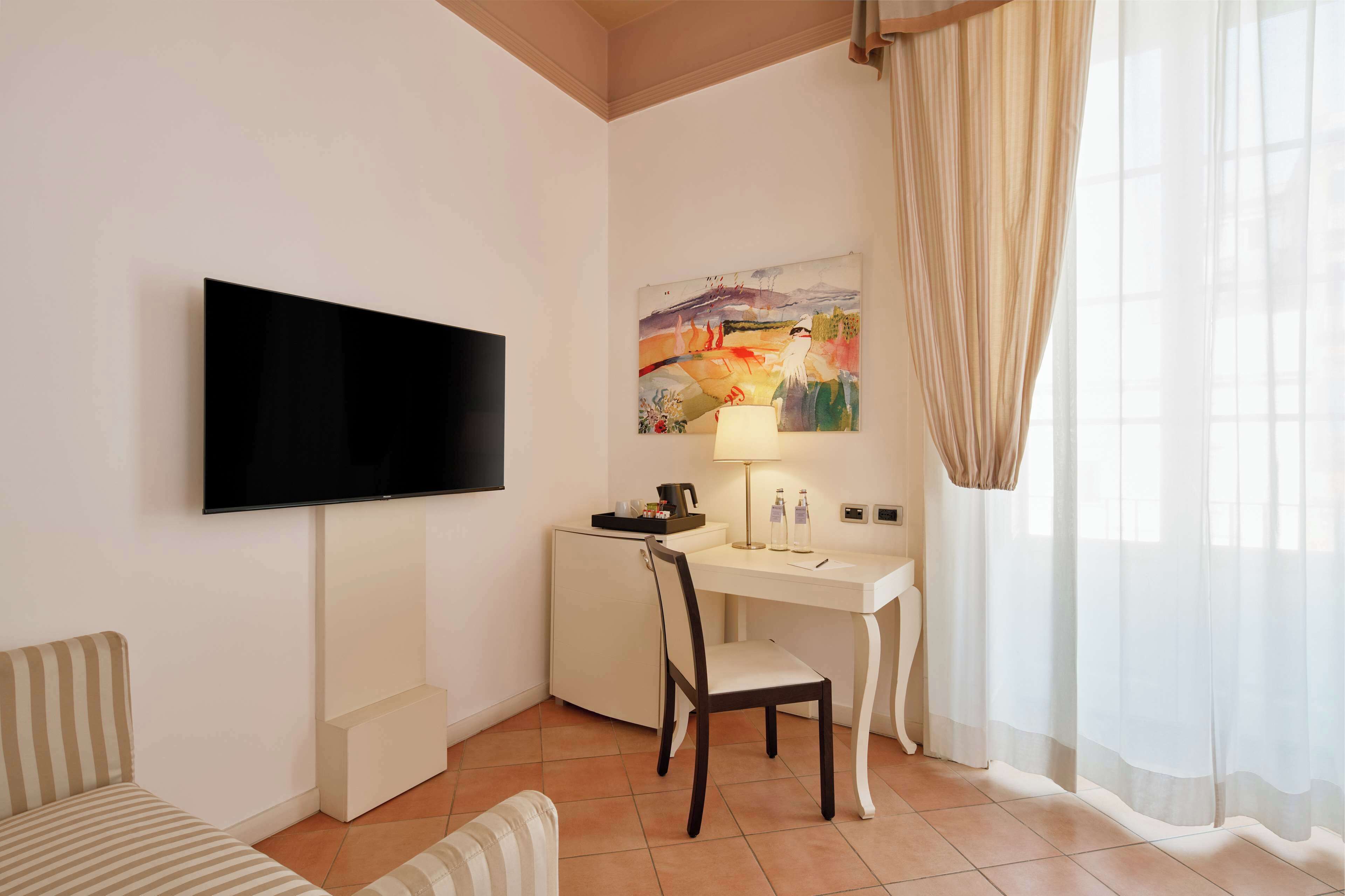 Quarto de hotel com televisão de tela plana na parede, uma escrivaninha com cadeira, pequeno frigorífico, abajur e um quadro na parede.