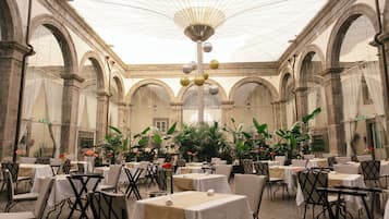 Uma área de refeições interna com mesas e cadeiras, um lustre decorativo central e arcos nas paredes.