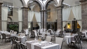 Um espaço de refeições interno com várias mesas e cadeiras, grandes janelas com cortinas e colunas de pedra.