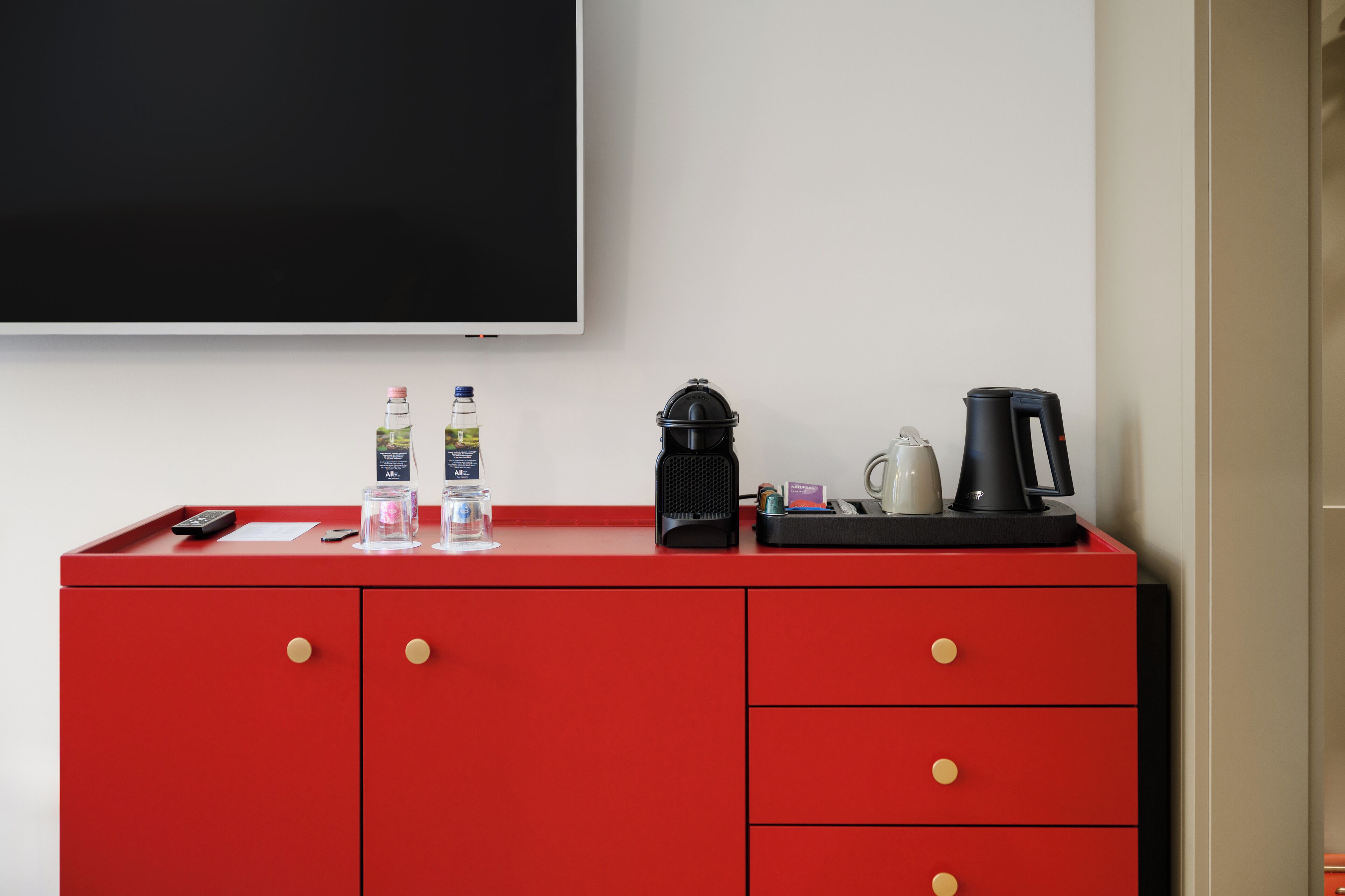 Um minibisco vermelho com um controle remoto, duas garrafas de água, uma máquina de café e um bule de chá.