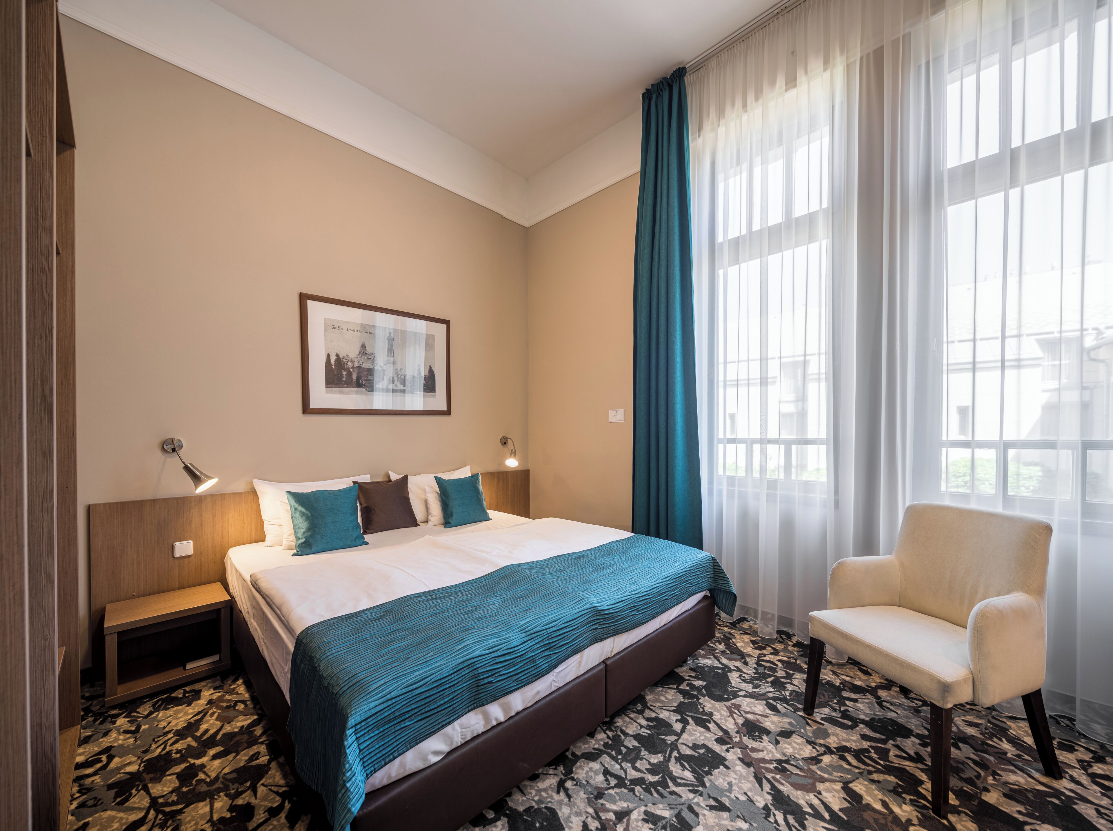 Quarto de hotel com uma cama grande, uma cadeira, uma mesinha de cabeceira, um abajur e um quadro na parede.